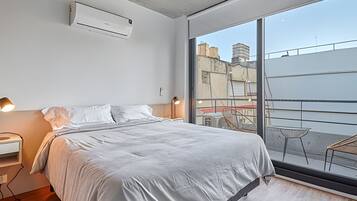 Quarto com uma cama grande, mesa de cabeceira, luminária e vista da cidade através de uma varanda.