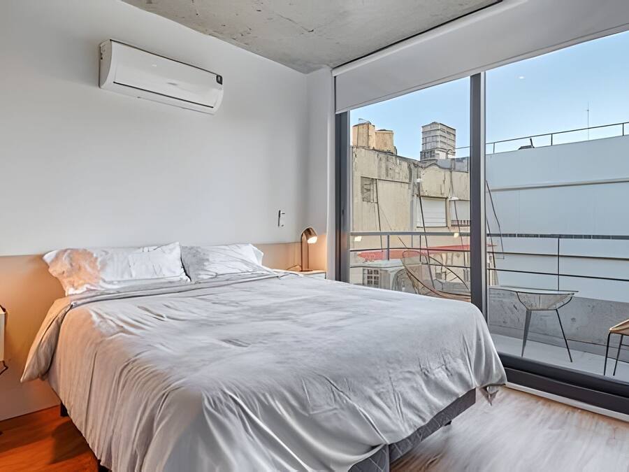 Quarto com uma cama grande, mesa de cabeceira, luminária e vista da cidade através de uma varanda.