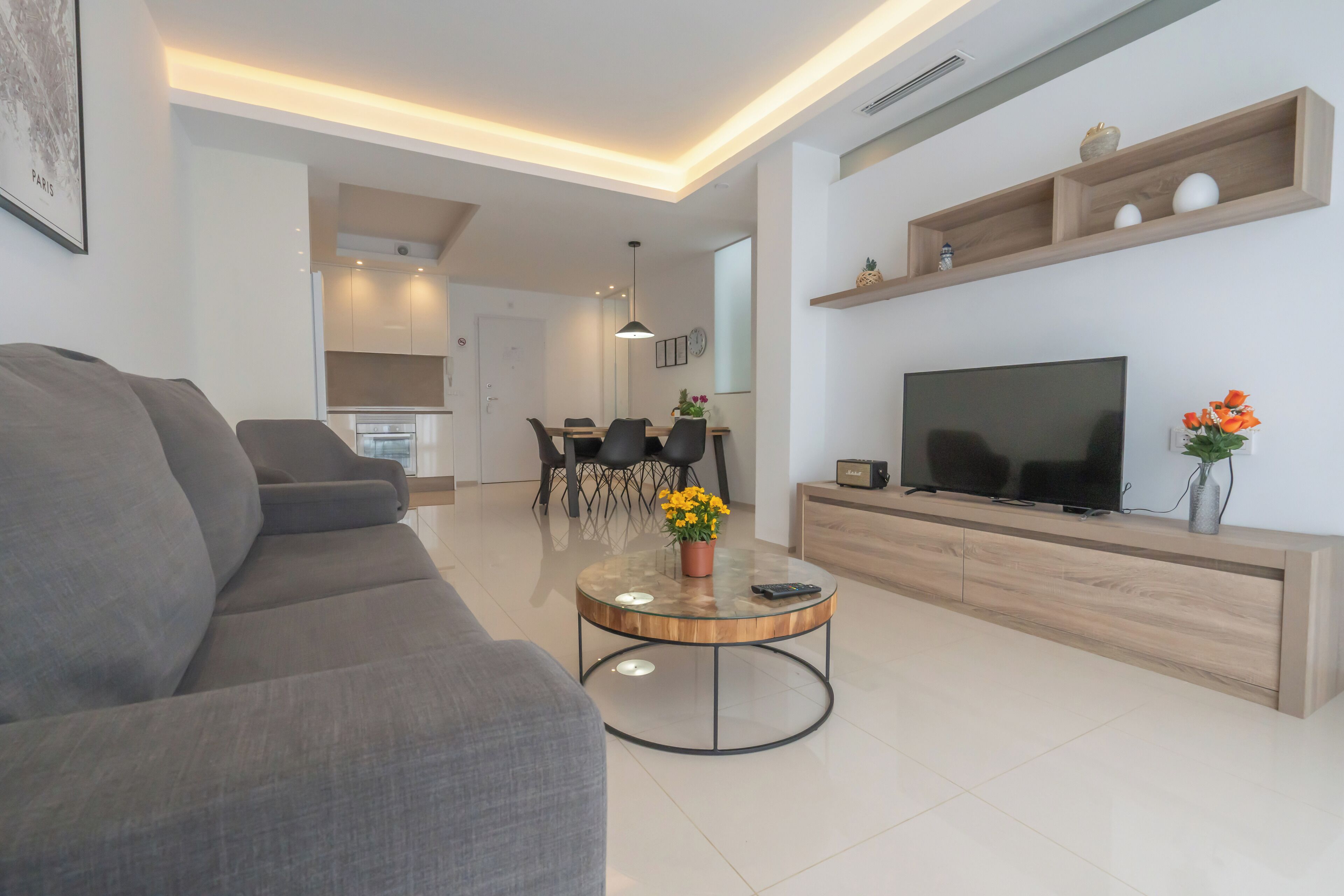 Uma sala de estar moderna com um sofá cinza, uma mesa de centro com um vaso de flores, uma TV sobre um suporte de madeira e uma cozinha ao fundo.
