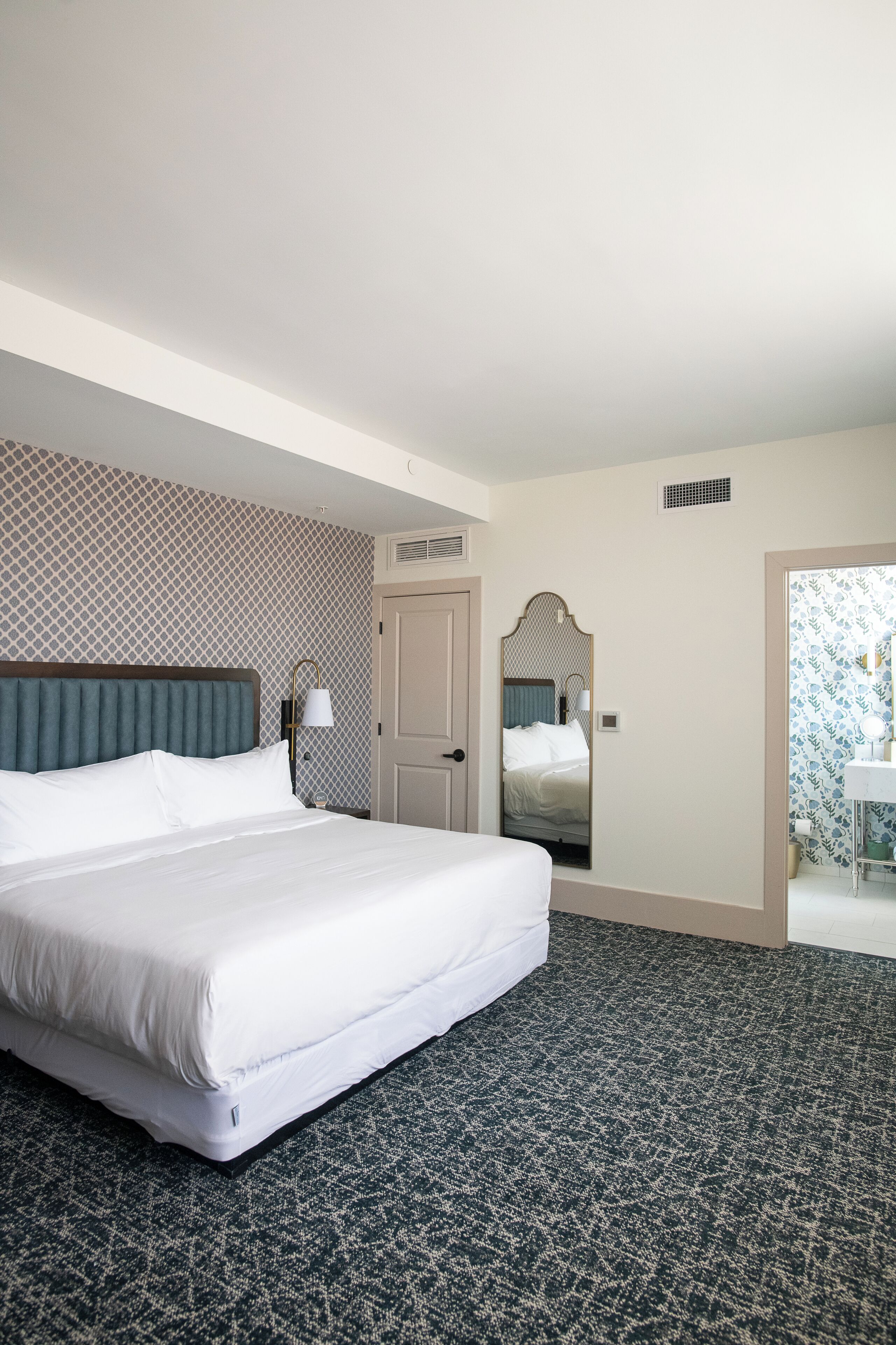 Quarto de hotel com uma cama grande, uma mesa de cabeceira com abajur, uma porta, um espelho e um banheiro visível através de uma porta aberta.