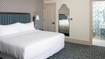 Quarto de hotel com uma cama grande, uma mesa de cabeceira com abajur, uma porta, um espelho e um banheiro visível através de uma porta aberta.