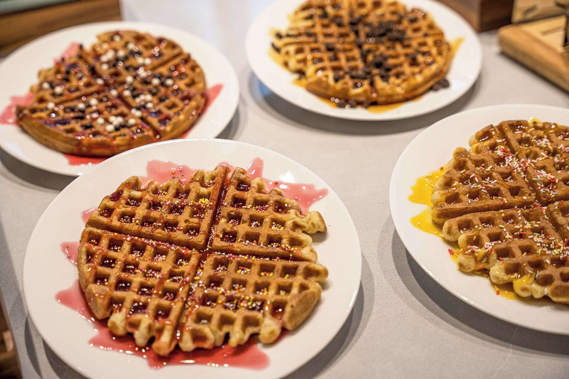 Quatro pratos de waffles com coberturas variadas.