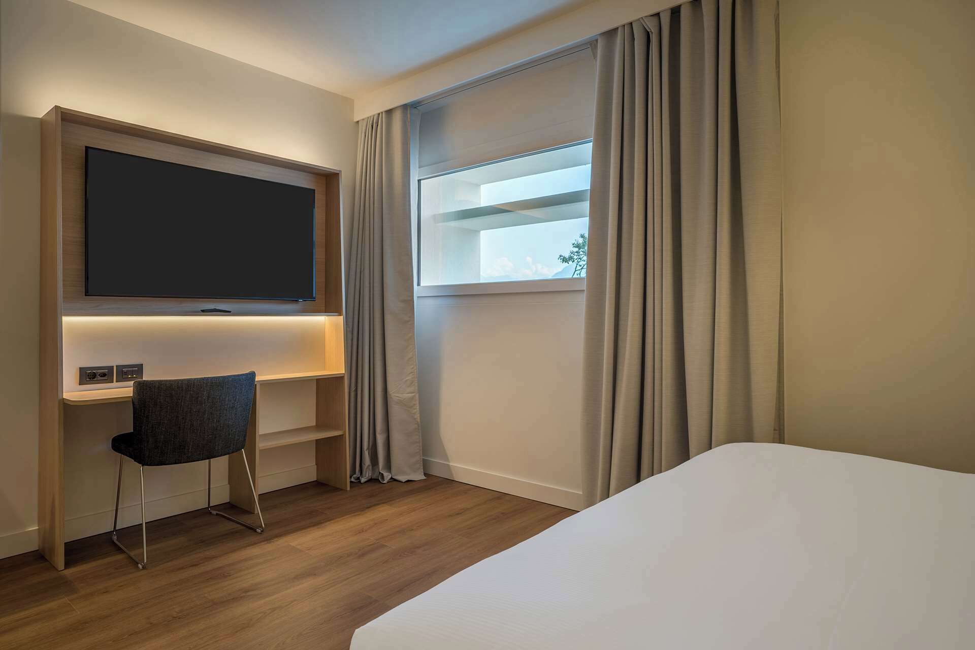 Quarto de hotel com uma cama, uma escrivaninha com cadeira, uma televisão na parede e uma janela com cortinas.