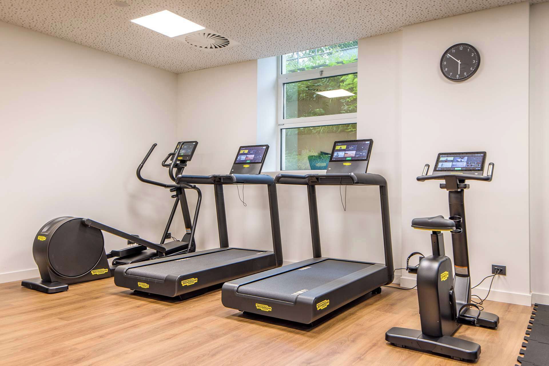 Sala de ginástica com três esteiras, um elíptico e uma bicicleta ergométrica, todos com mostradores digitais.