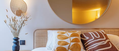Uma cama bem arrumada com roupa de cama branca e almofadas decorativas, um espelho redondo na parede e um vaso com plantas secas em uma mesa de cabeceira.