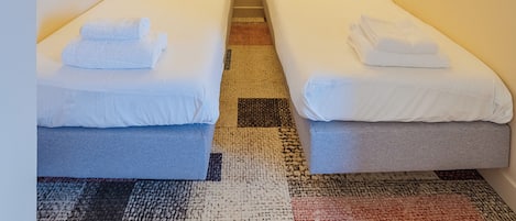 Quarto pequeno e bem iluminado com duas camas de solteiro, cada uma com travesseiro e toalha dobrada. Há um espelho redondo na parede e um tapete com textura no chão.