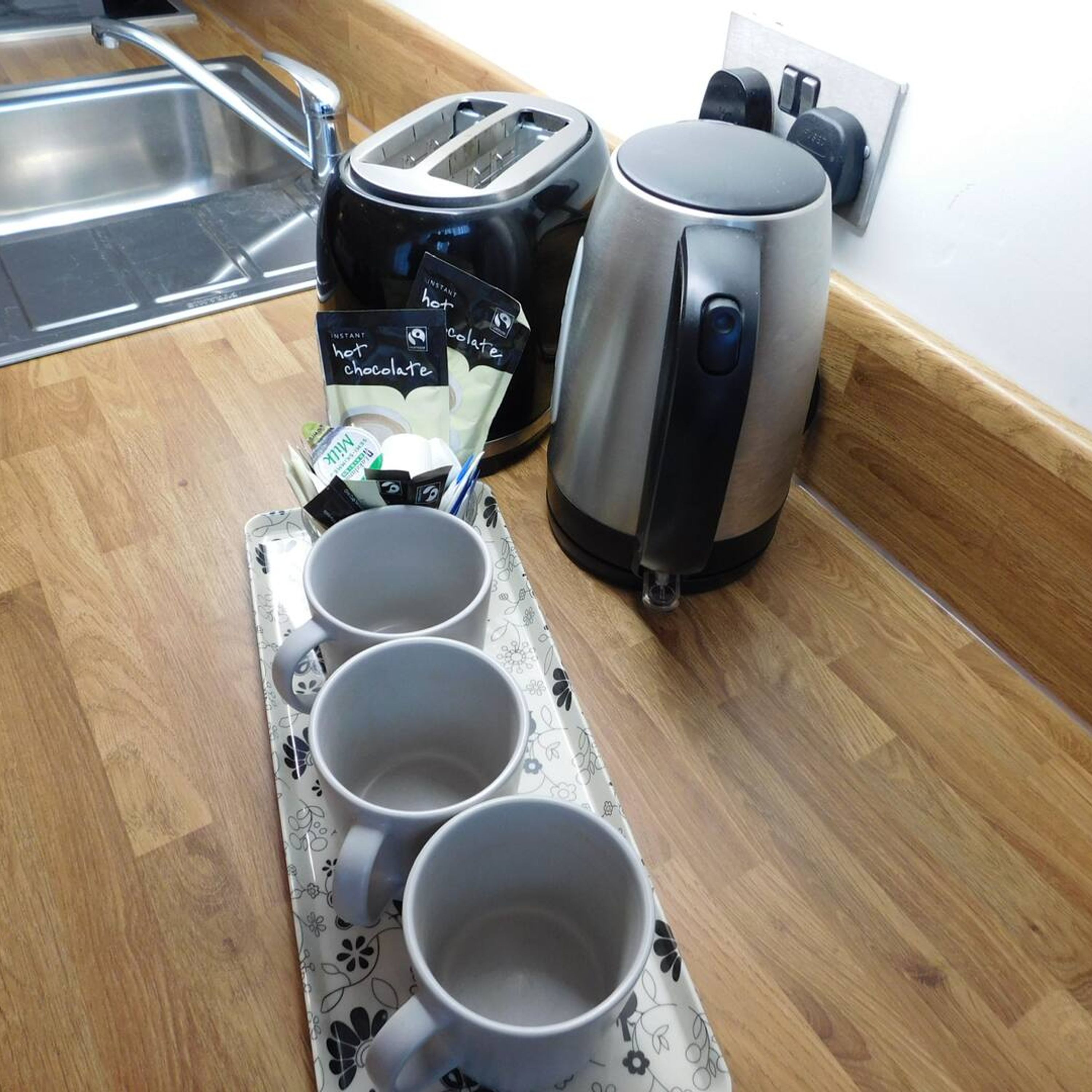 Uma bancada de cozinha com uma torradeira, uma cafeteira e quatro chávenas numa bandeja estampada com flores.