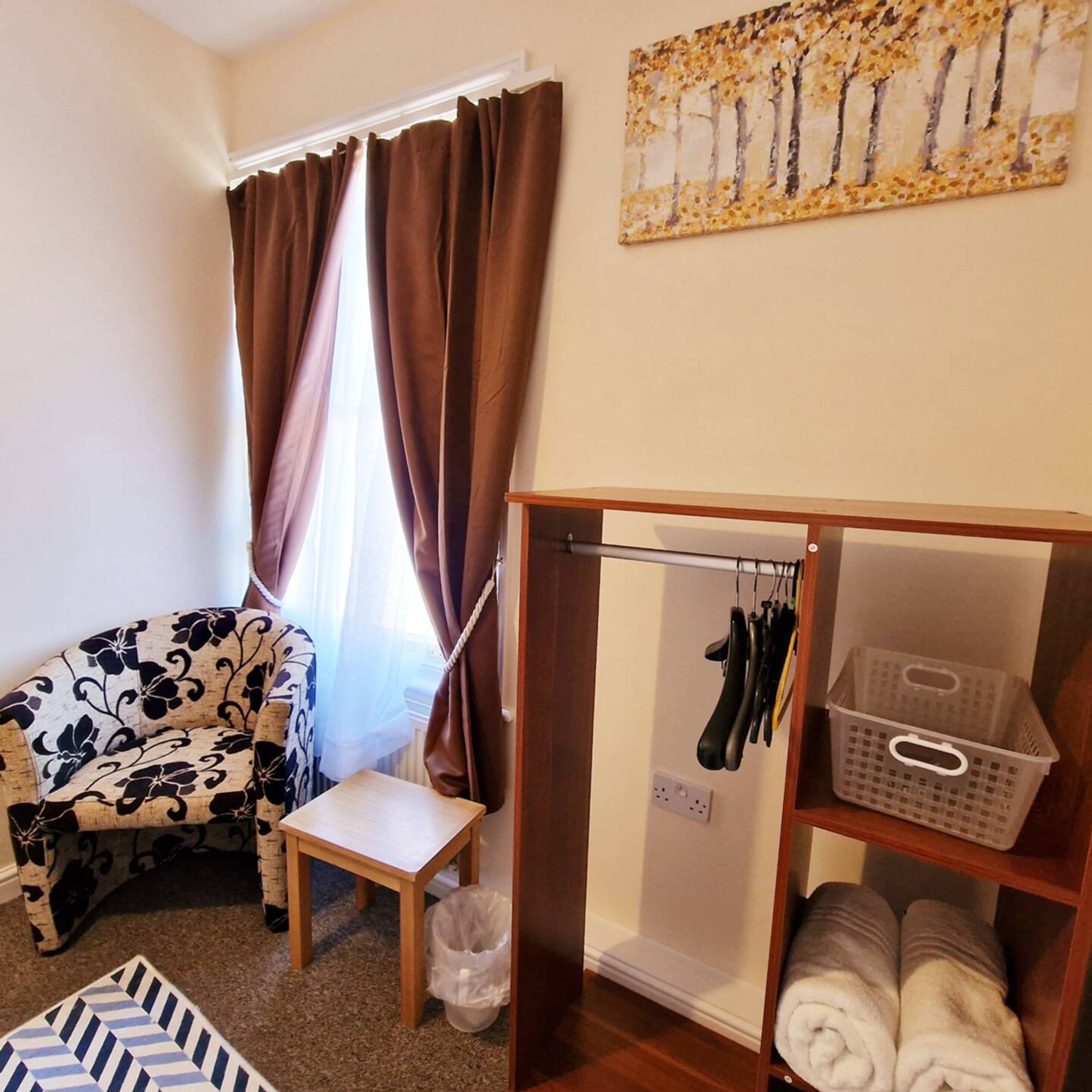 Um quarto pequeno com uma poltrona estofada, uma prateleira de madeira com cestos de armazenamento e um quadro na parede.