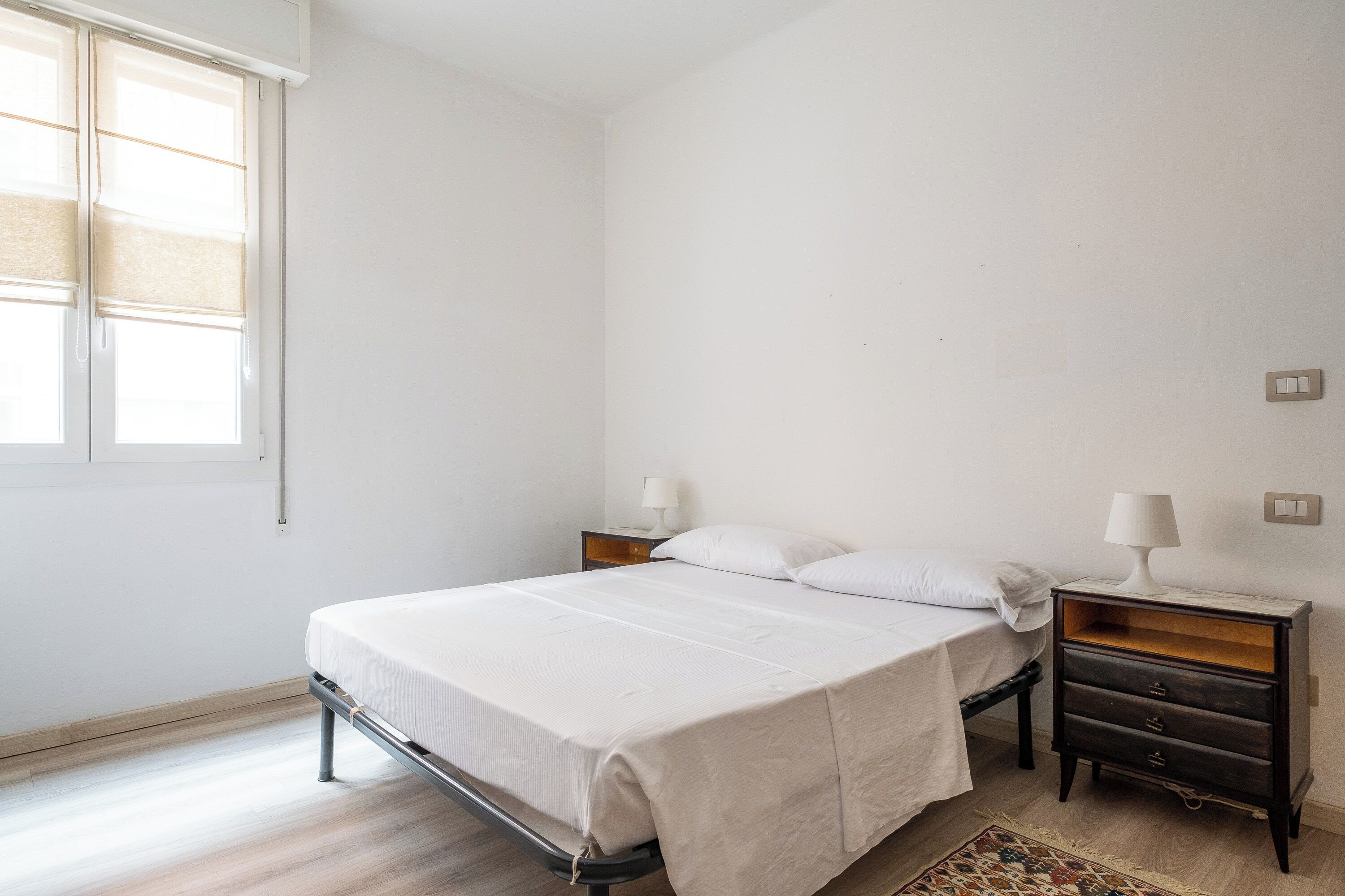 Quarto com cama, duas mesas de cabeceira com abajures, janela com persianas e piso de madeira.