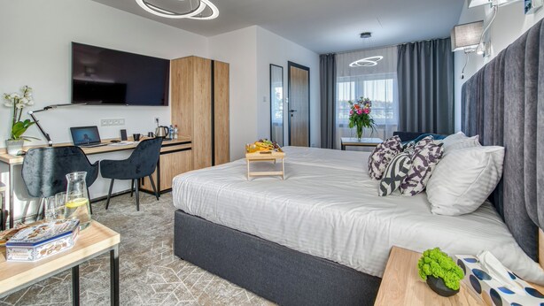 Quarto de hotel moderno com uma cama grande, uma escrivaninha com um laptop, uma televisão e vista para a cidade.