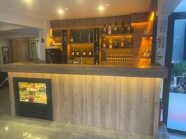 Uma área de bar com prateleiras de madeira exibindo garrafas de bebidas e copos, um refrigerador com sobremesas e um extintor de incêndio no canto.