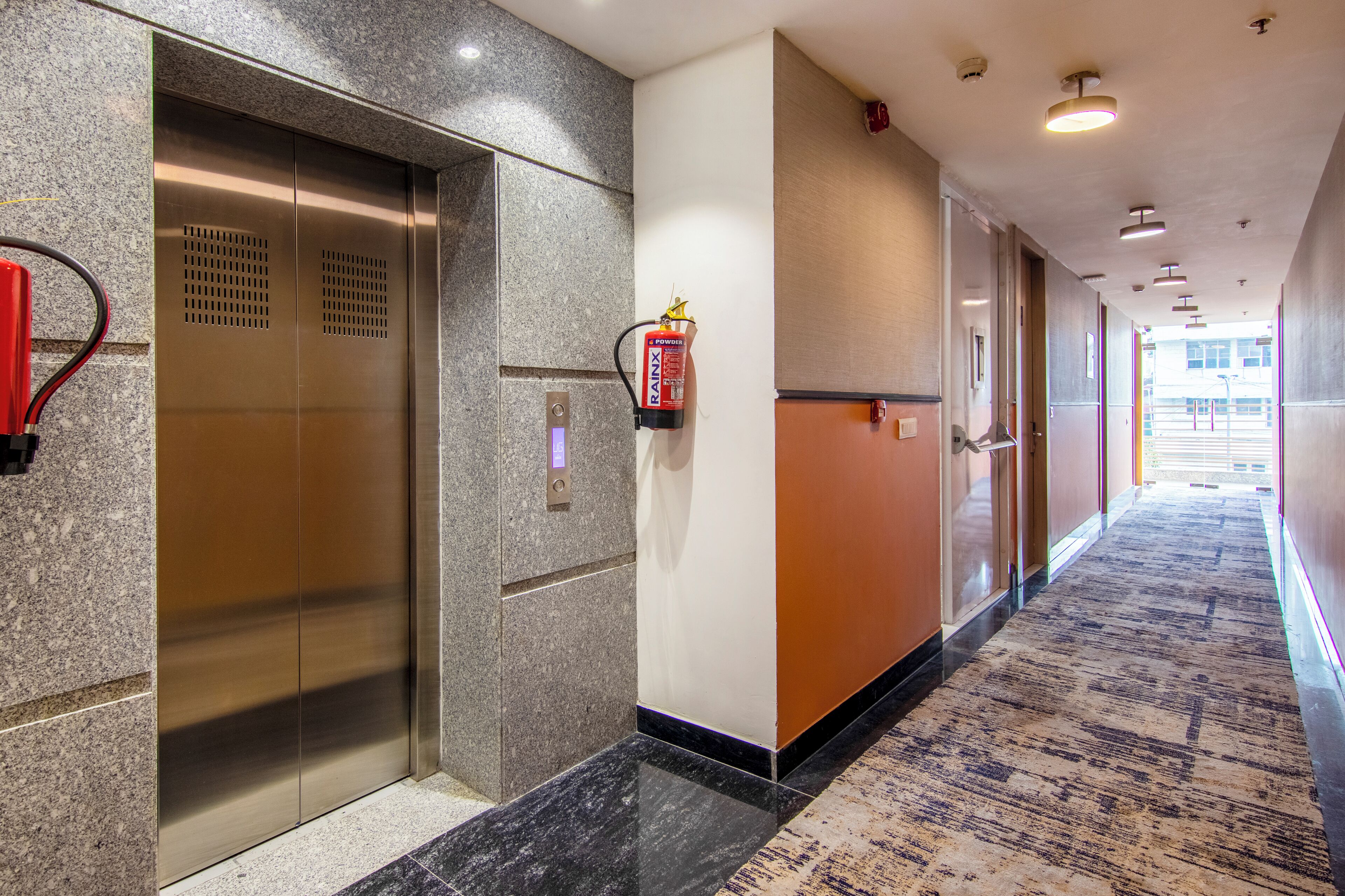 Corredor de hotel com elevador, extintor de incêndio e piso carpetado.