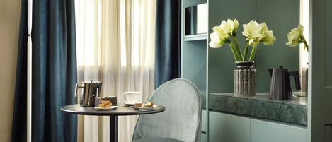 Quarto moderno com uma poltrona estofada cinza, uma pequena mesa redonda com uma xícara e um prato de comida, um vaso com flores e um armário verde com espelho.