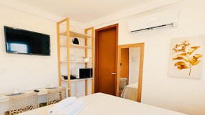 Quarto de hotel com cama, televisão, micro-ondas, prateleira com utensílios de cozinha, porta e um quadro na parede.