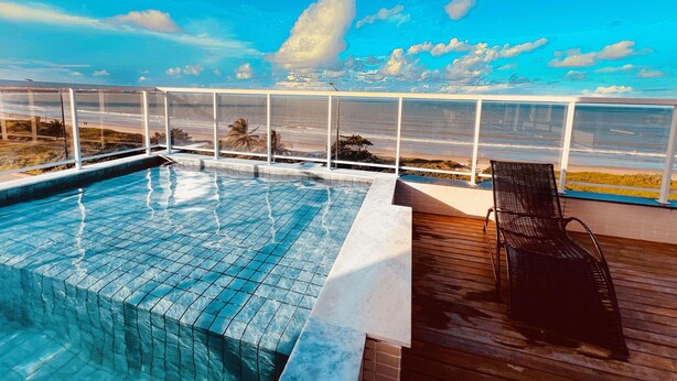 Piscina com vista para a praia, área de deck coberta e uma cadeira.