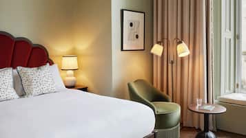 Quarto de hotel com uma cama, uma poltrona verde, uma mesinha com um copo e um abajur.