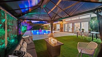 Um pátio externo com estrutura de vidro, piscina e área de refeições com cadeiras e mesa.