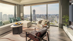 Quarto de hotel moderno com uma janela ampla que oferece vista para a cidade, um sofá, uma mesa de centro e uma poltrona de couro.