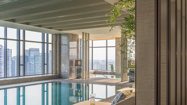 Piscina interior moderna com vista para a cidade, grandes janelas e área de estar com mesa de vidro e almofadas.