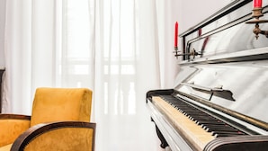 Um piano, uma vela vermelha, uma cadeira amarela e uma janela com cortinas translúcidas.