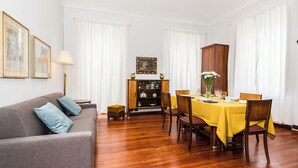 Uma sala de jantar com mesa de madeira posta para quatro pessoas, um sofá cinza com almofadas azuis e um armário com objetos decorativos.