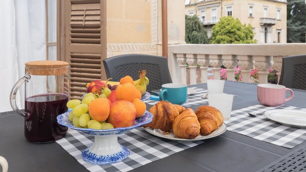 Uma mesa com um jogo americano xadrez, um jarro de bebida vermelha, uma fruteira com uvas e laranjas, croissants e uma caneca rosa.