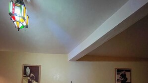 Quarto com mesa de madeira, duas cadeiras de vime, uma cama e duas fotos emolduradas nas paredes.
