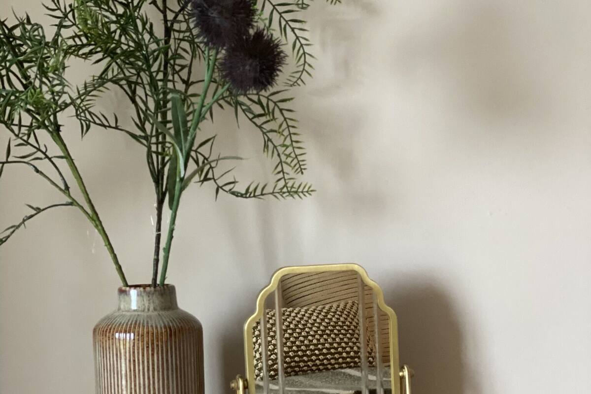 Uma mesa de madeira redonda com um vaso alto e canelado contendo folhagem verde e flores roxas, um pequeno pote de cerâmica branca e uma cadeira de arame com moldura dourada.