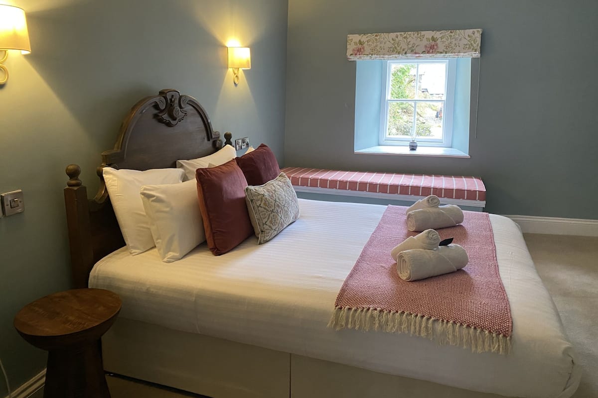 Quarto com cama, mesa de cabeceira, abajur fixado na parede, janela com cortina florida e vista para o verde lá fora.
