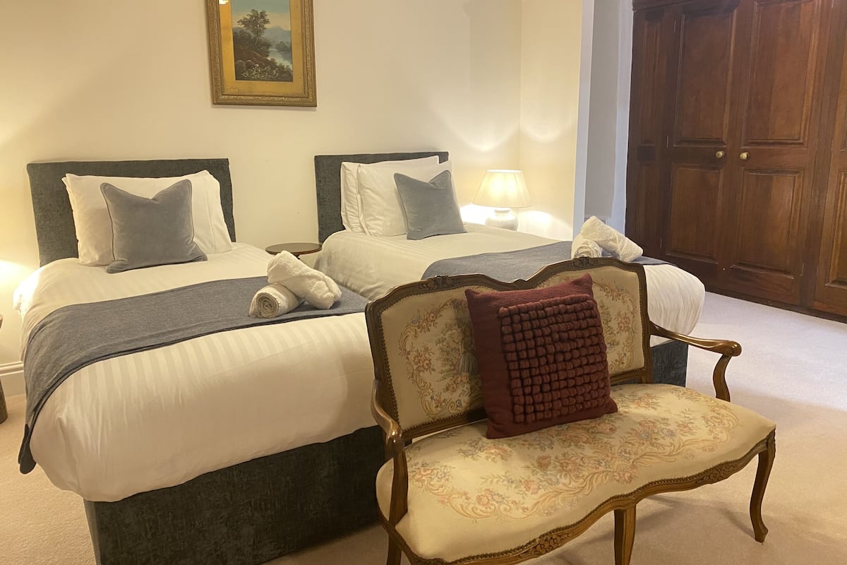 Quarto de hotel com duas camas, uma cadeira, um abajur e uma porta de madeira.