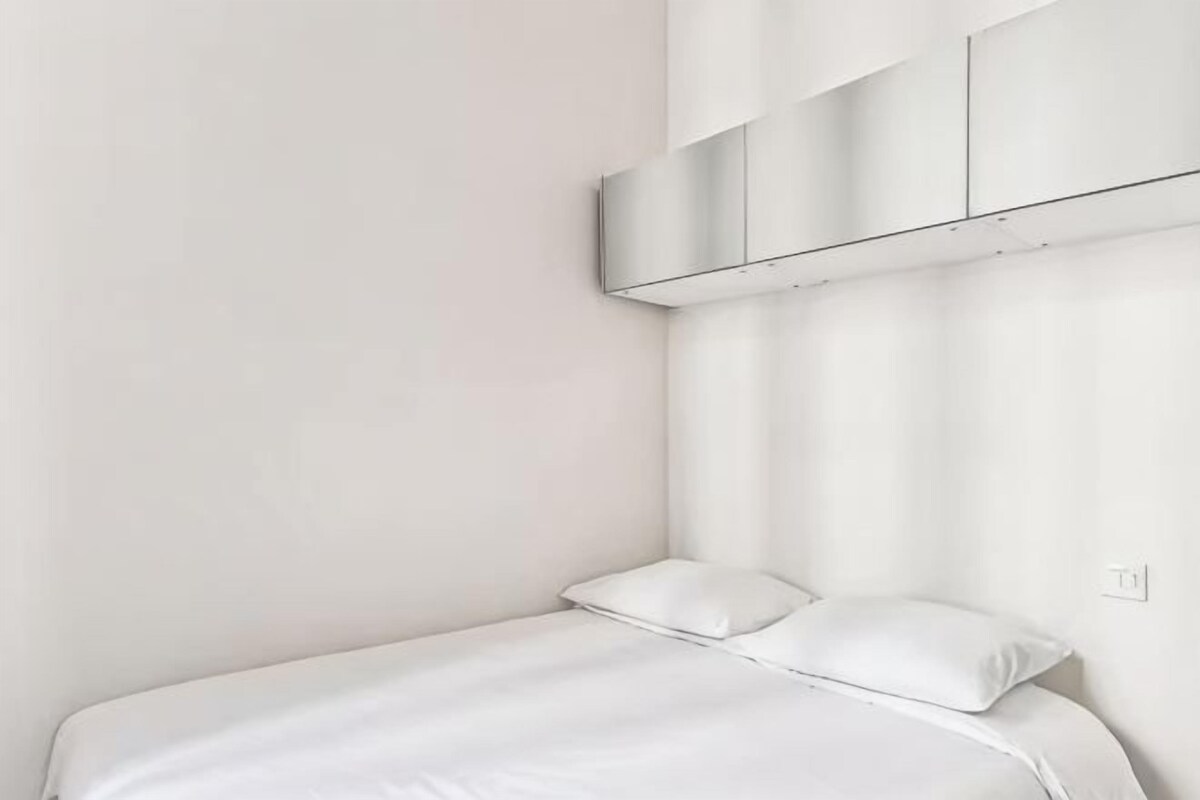 Cama branca com duas almofadas, parede branca e teto branco.