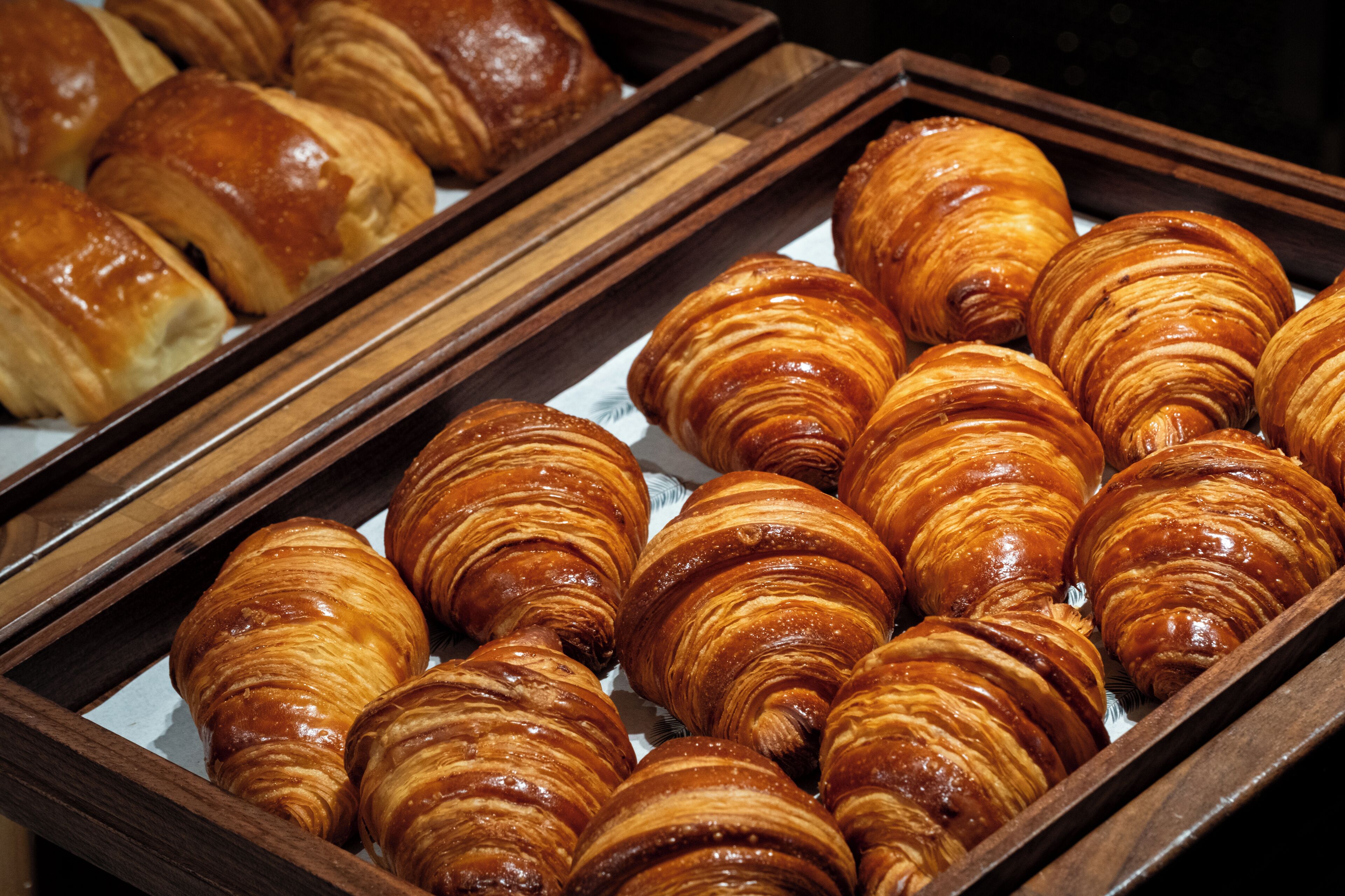 Exposição de diversos doces e pastelaria, incluindo croissants e outros produtos de panificação, dispostos em tabuleiros de madeira.