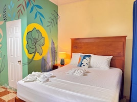 Cama bem arrumada com lençóis brancos, cabeceira de madeira e um mural colorido com motivos florais.