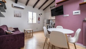 Uma sala de estar moderna com um sofá roxo, cadeiras brancas, uma mesa de madeira e uma área de cozinha com televisão e ventoinha.