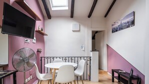 Uma área de refeições com uma mesa redonda, cadeiras brancas, um ventilador e uma TV fixada na parede.