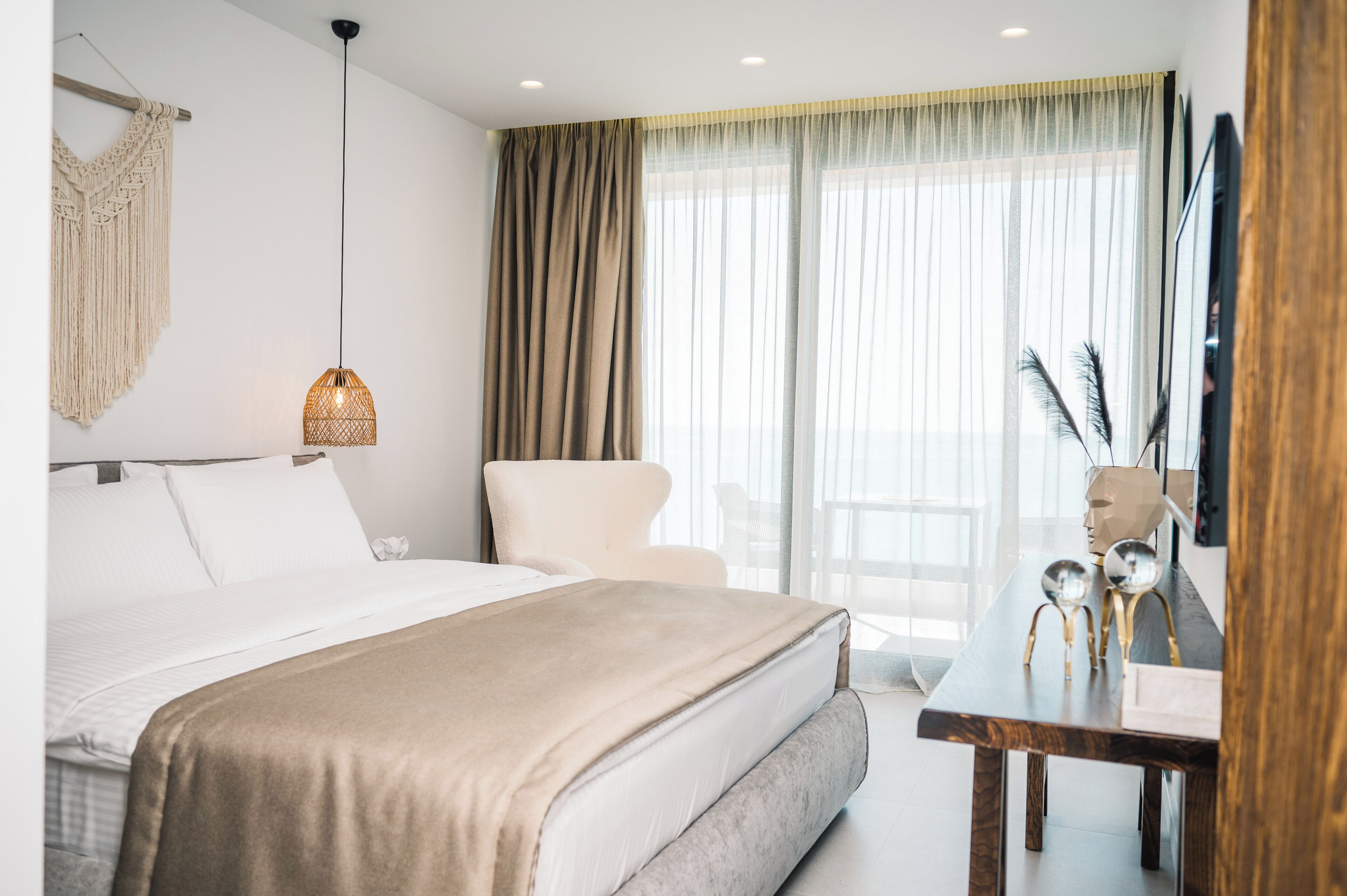 Quarto de hotel moderno com uma cama grande, uma escrivaninha com abajur e vista para o exterior através de cortinas leves.