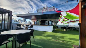 Uma área externa com mesas e cadeiras, um bar chamado 'Fel RooTop' e vista para o horizonte da cidade.