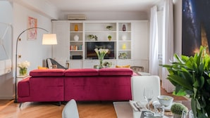 Uma sala de estar moderna com um sofá vermelho, uma TV de tela plana, prateleiras brancas e uma janela grande com cortinas.