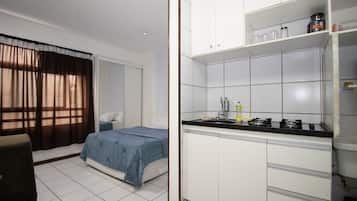 Quarto compacto com cama, pequena mesa de escritório, cozinha compacta equipada com fogão e pia, e janela com cortinas.