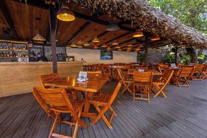 Um restaurante ao ar livre com mesas e cadeiras de madeira, um espaço de bar com garrafas, e um telhado de palha.