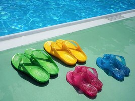 Uma área à beira da piscina com superfície verde e quatro pares de chinelos nas cores verde, amarelo, rosa e azul.