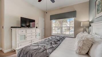 Um quarto com uma cama, uma televisão sobre um penteadeira, uma janela com persianas e um ventilador de teto.