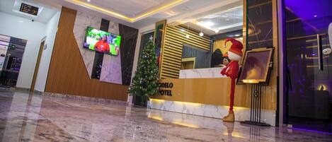 Um hall de hotel com uma mesa de recepção, um pinheiro de Natal e uma grande tela de televisão.