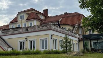 Uma casa grande de dois andares com telhado de telhas vermelhas, várias janelas e uma varanda com balaustrada.