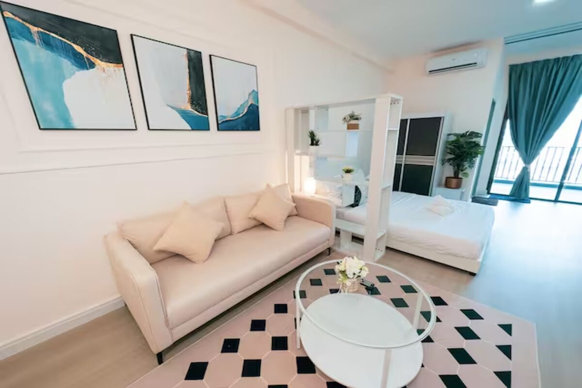 Quarto de hotel moderno com um sofá bege, uma mesa branca redonda e uma cama. Há três obras de arte emolduradas na parede, uma planta em vaso e uma janela com cortinas.