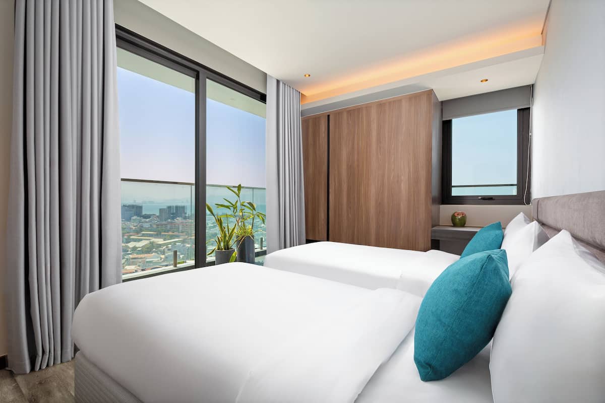 Quarto de hotel com duas camas, uma janela grande com vista para a cidade, um guarda-roupa de madeira e uma pequena mesa com um vaso.