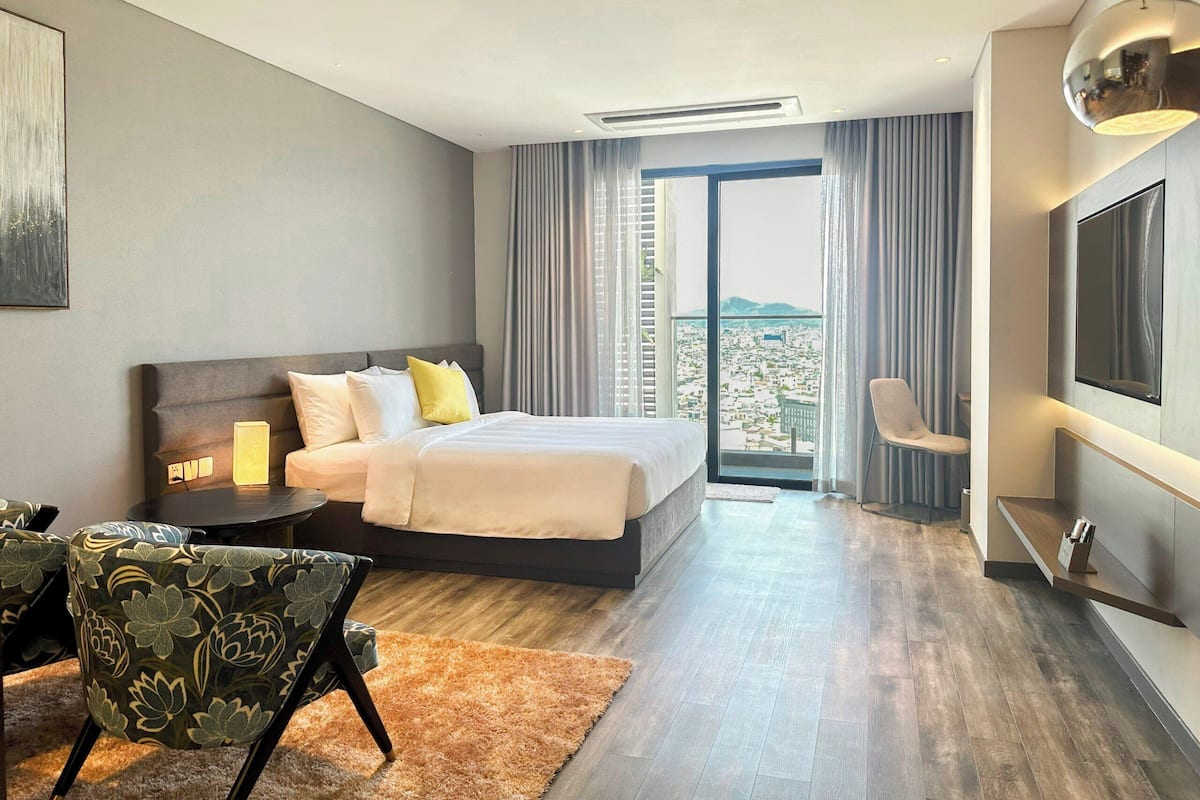 Quarto de hotel moderno com uma cama grande, televisão na parede, varanda com vista para a cidade e área de estar com uma cadeira e mesa.