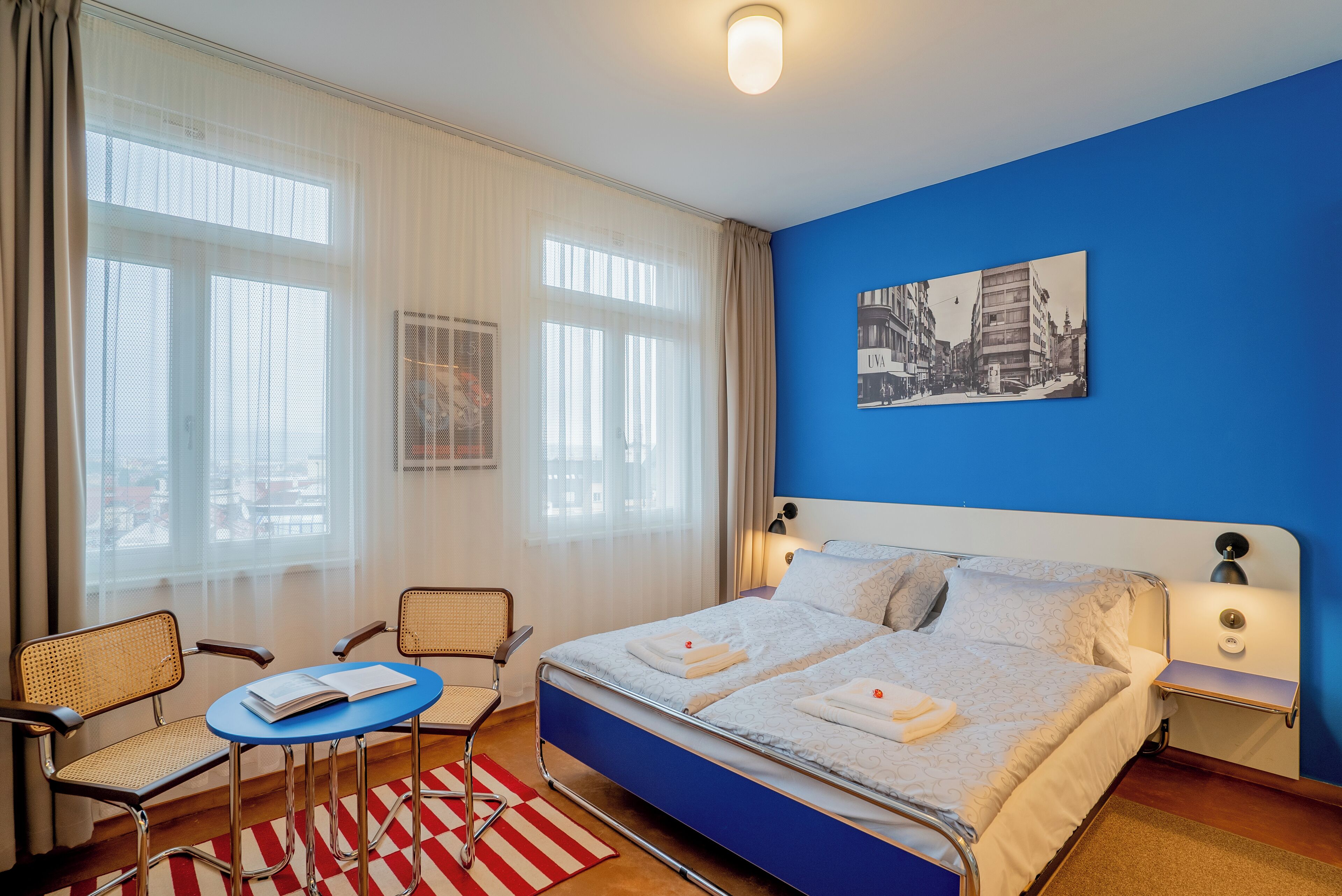 Quarto de hotel com cama, duas cadeiras, uma mesa pequena e uma janela com cortinas.