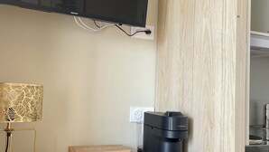Quarto moderno com televisão de tela plana na parede, escrivaninha de madeira com luminária, cafeteira e uma caixa.