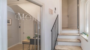 Uma escadaria com guarda-corpo de metal, uma porta com painel de vidro e uma pequena mesa com uma planta.